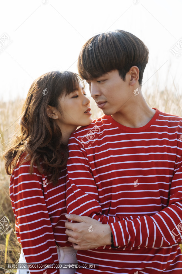 卷曲的长发女孩试图亲吻她的男朋友的脸颊,而他们站在一起在草原上。摄影素材汇图网